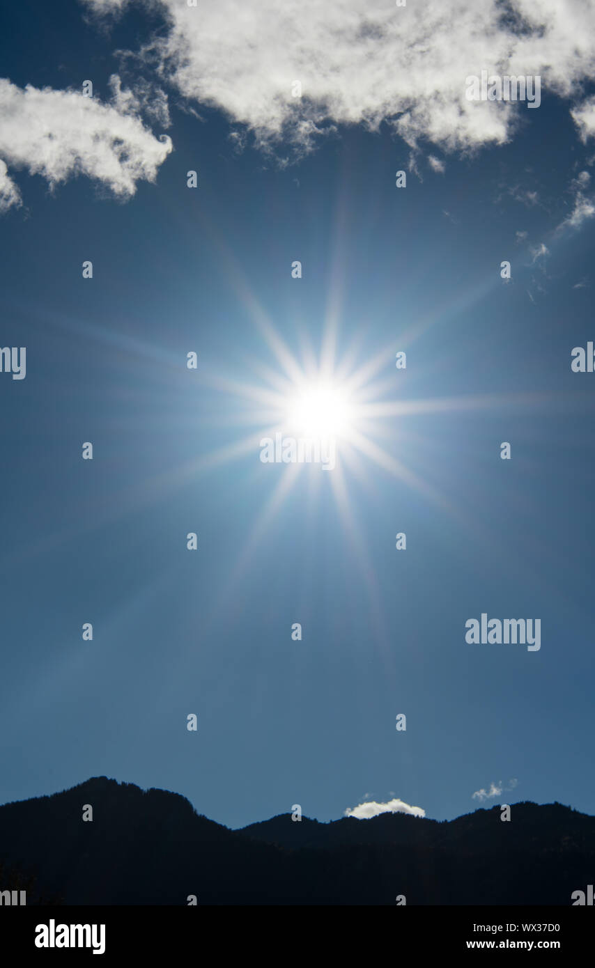 sun shines brightly in a star shape in the brigh blue sky with a few ...