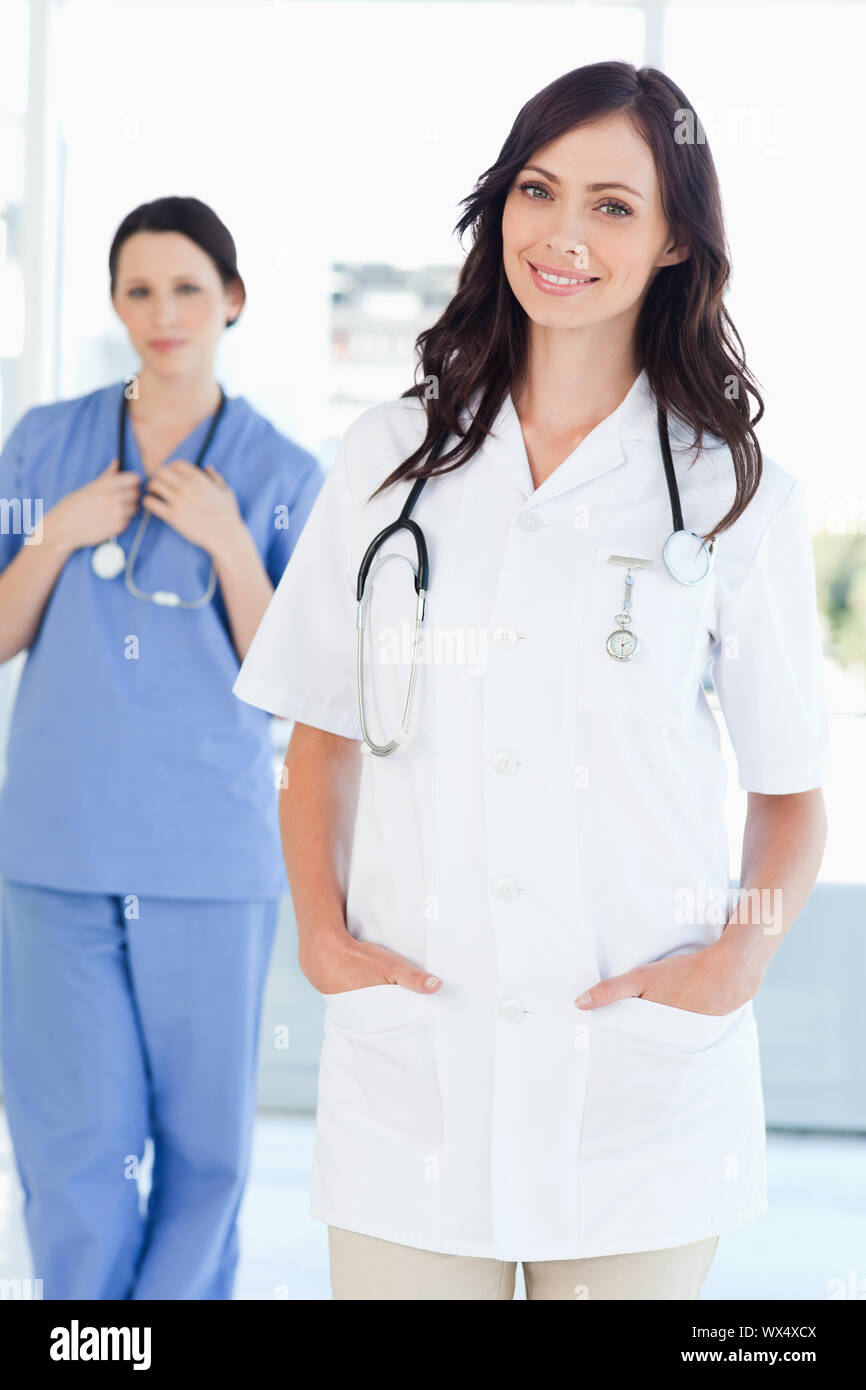 A smiling nurse standing upright while her colleague is behind her ...