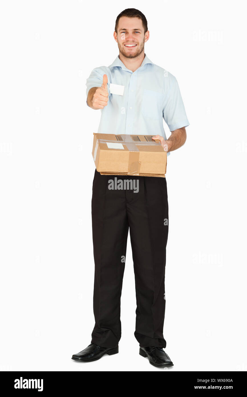 Smiling young post employee with parcel giving thumb up against a white ...