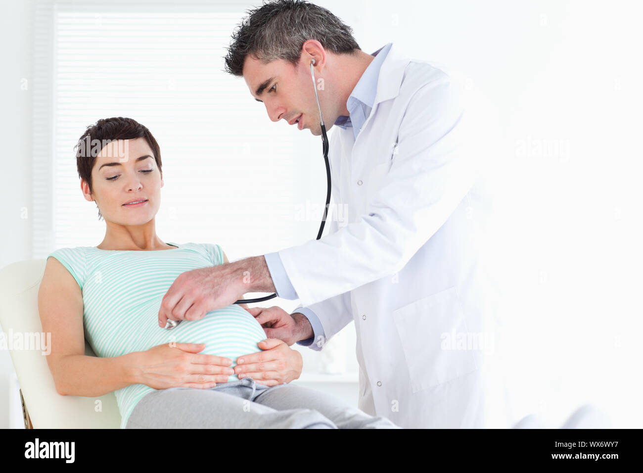 Doctor examining the cute woman's tummy with a stethoscope in a room ...