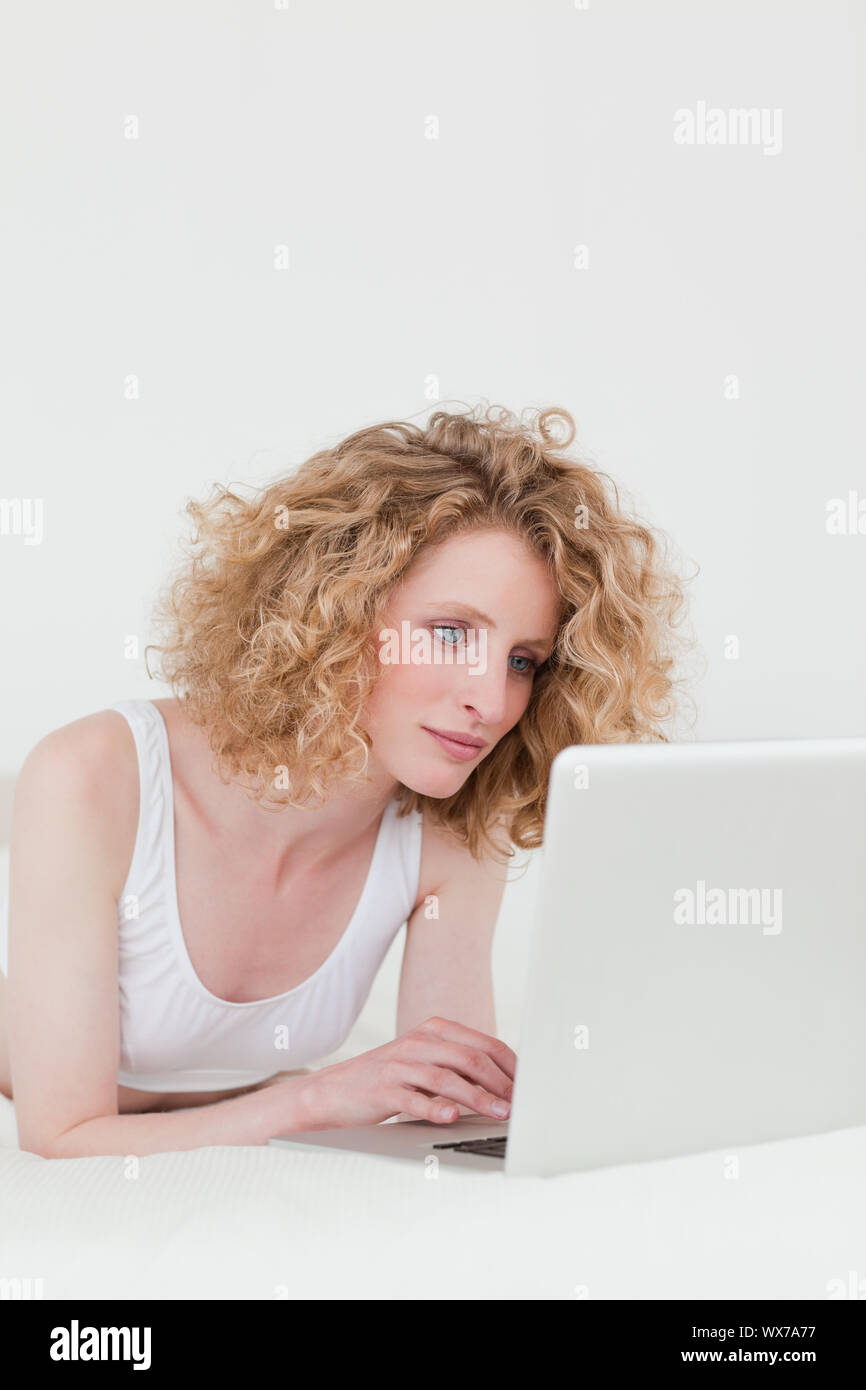 Cute blonde woman relaxing with her laptop while lying on her bed in ...