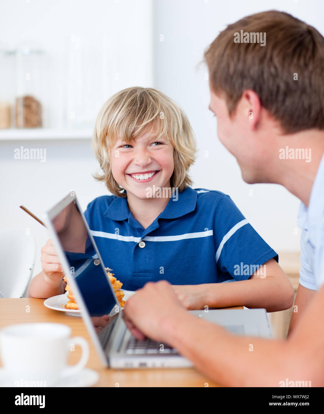 Smiling boy having breakfast while his father using a laptop in the ...