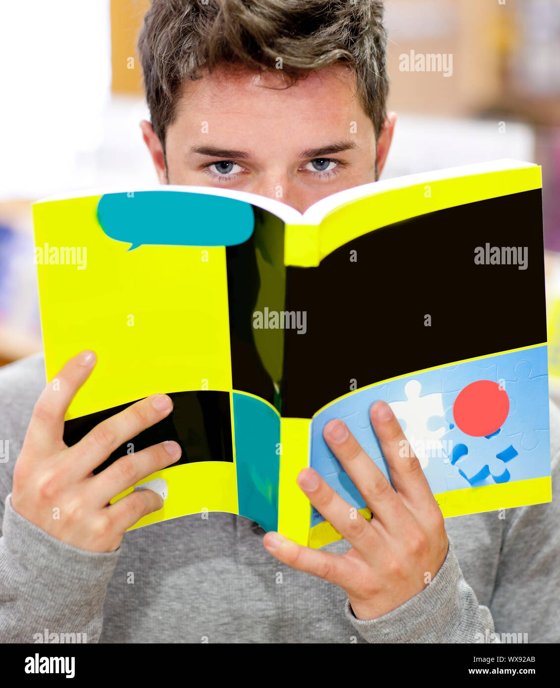 Male student standing in a book shop hiding his face behind a book ...