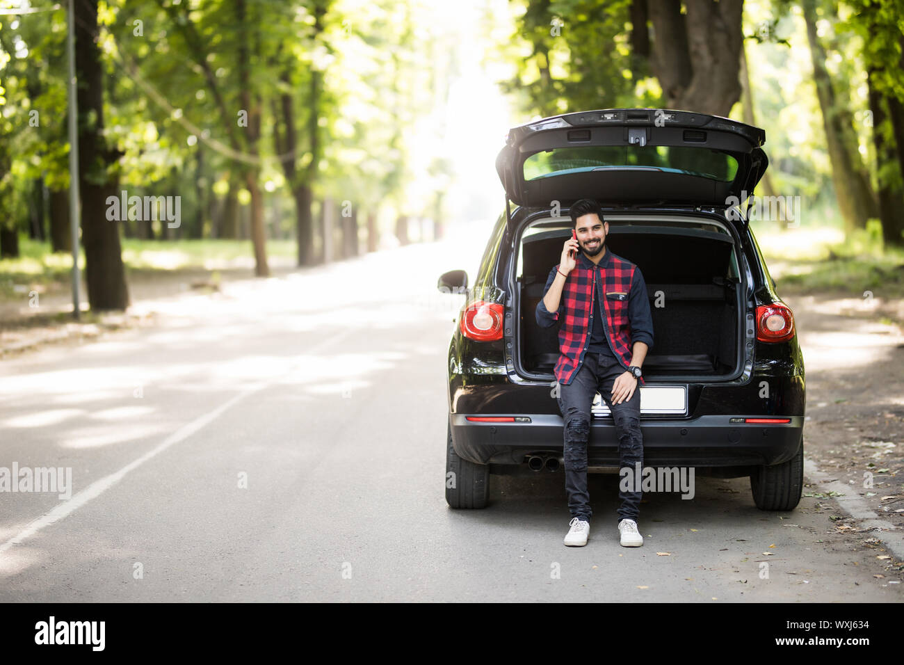 Young indian man sitting in the trunk of his car on the street road ...