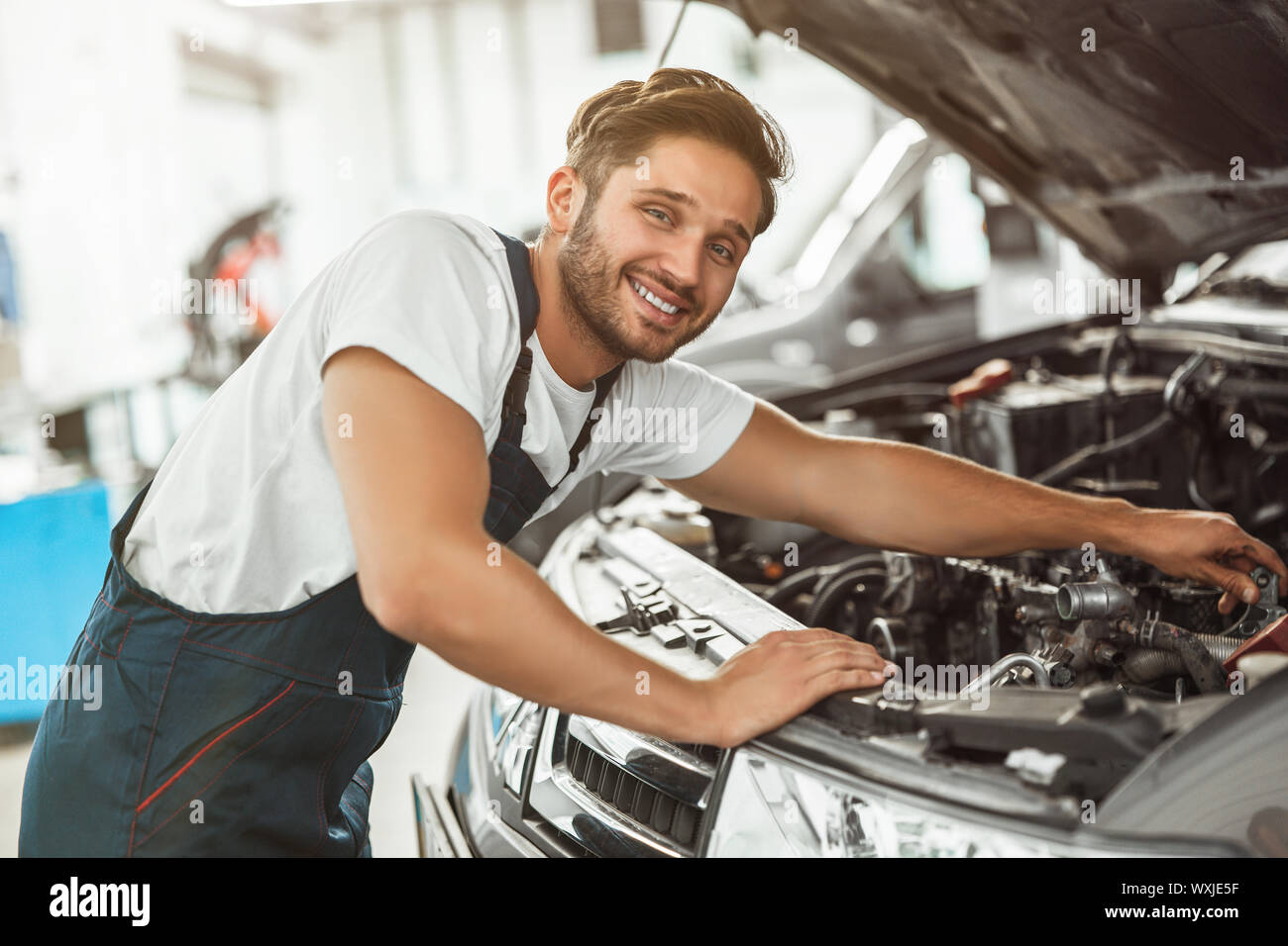 young smiling handsome mechanic in uniform fixing motor in car bonnet ...