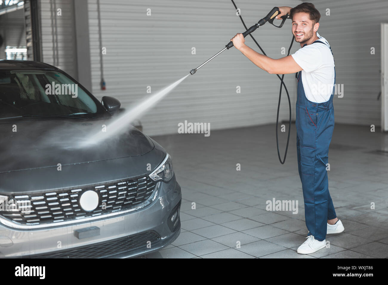 young handsome man wearing uniform washing car at car washing station ...