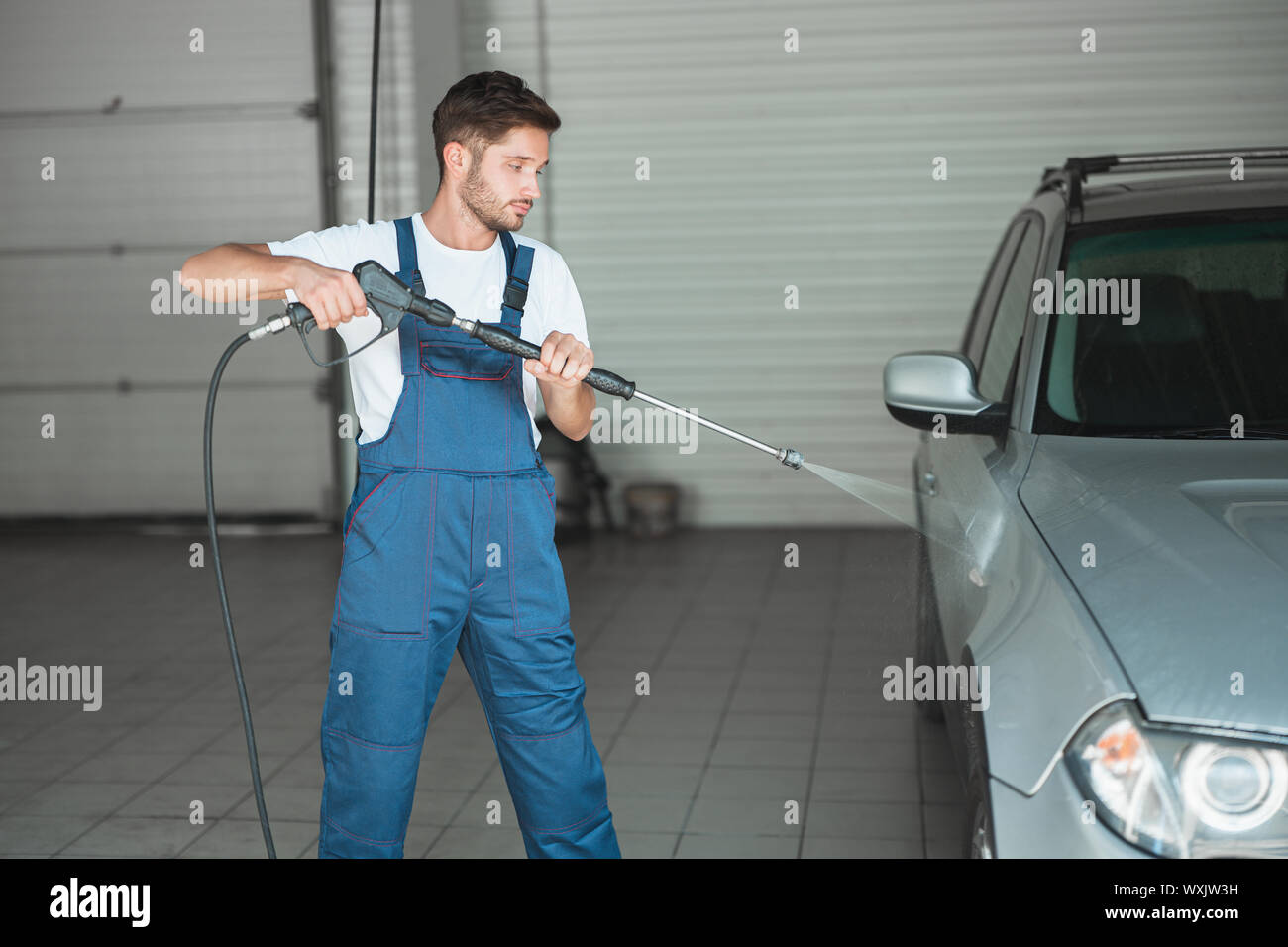 young handsome man wearing uniform washing car at car washing station ...