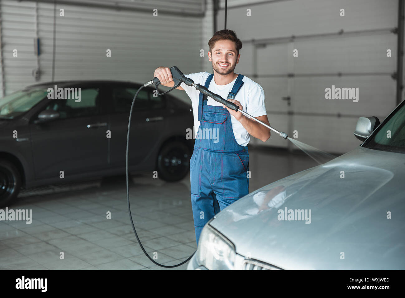 young handsome man wearing uniform washing car at car washing station ...