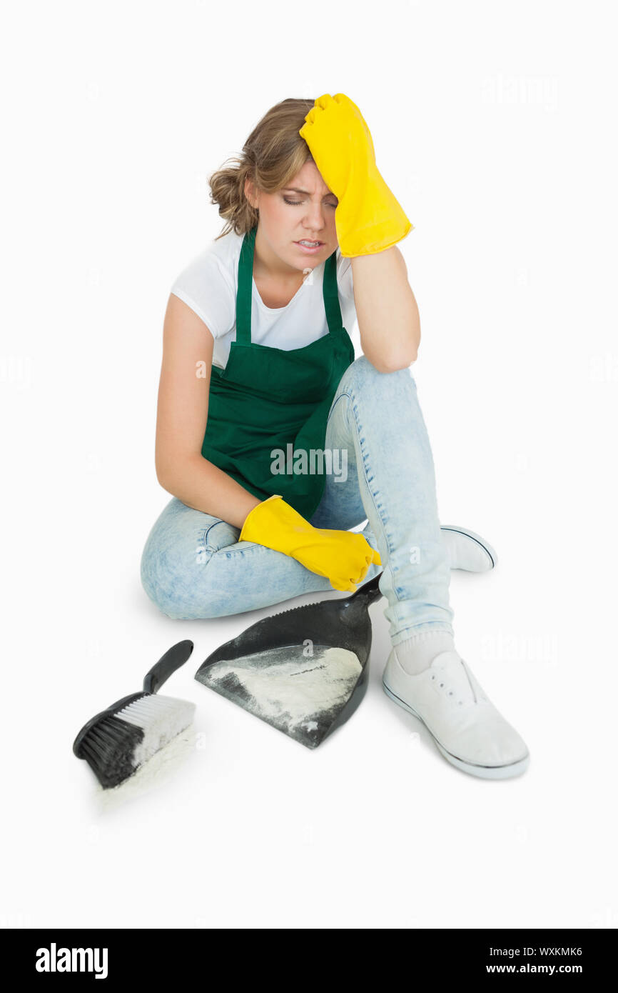 Tired young maid sitting with brush and dust pan over white background ...