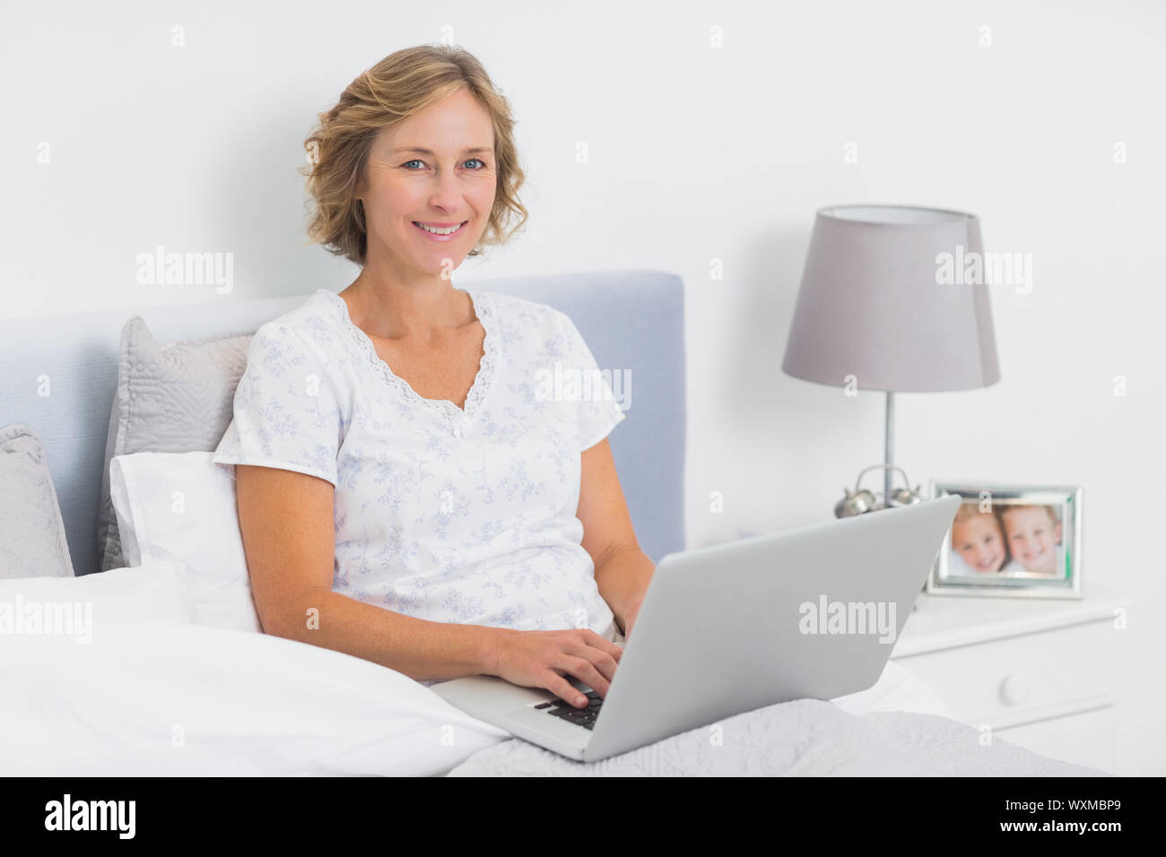 Content Blonde Woman Sitting In Bed Using Laptop Smiling At Camera At