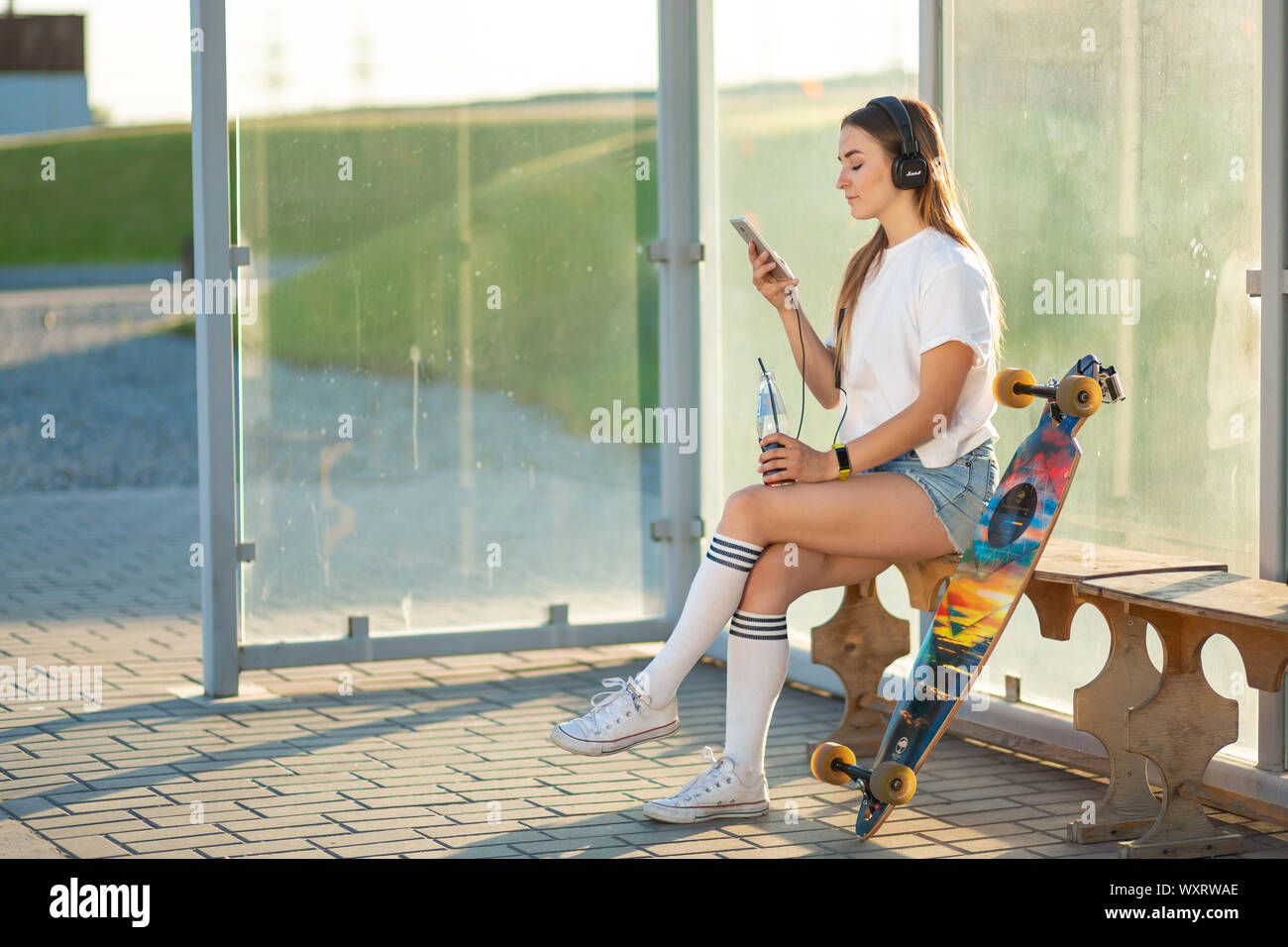 Stylish young girl sitting on bus stop with her longboard, listening ...