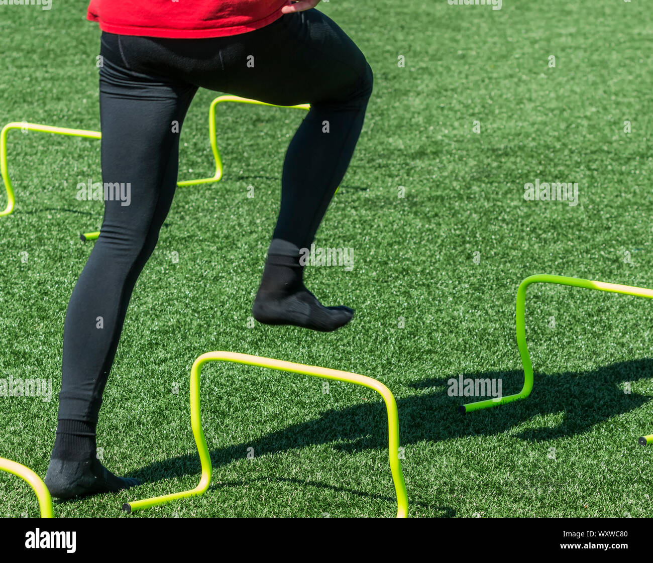 An athlete wearing a red shirt and black spandex is stepping over yellow mini banana hurdles ...