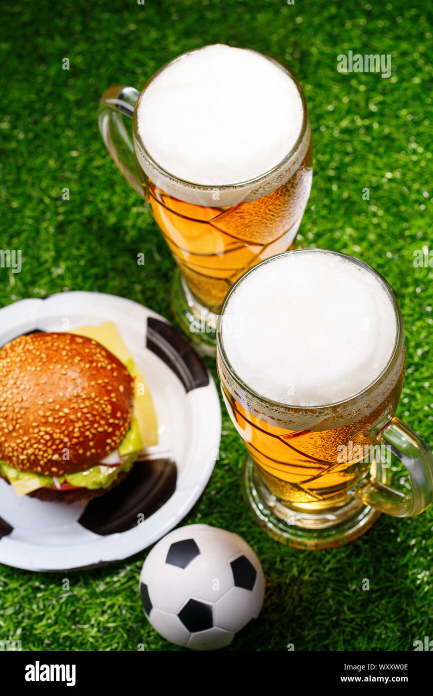 Two glasses of beer, hamburger and soccer ball on green grass. Table ...