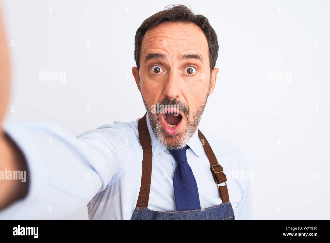 Middle age shopkeeper man wearing apron make selfie over isolated white ...