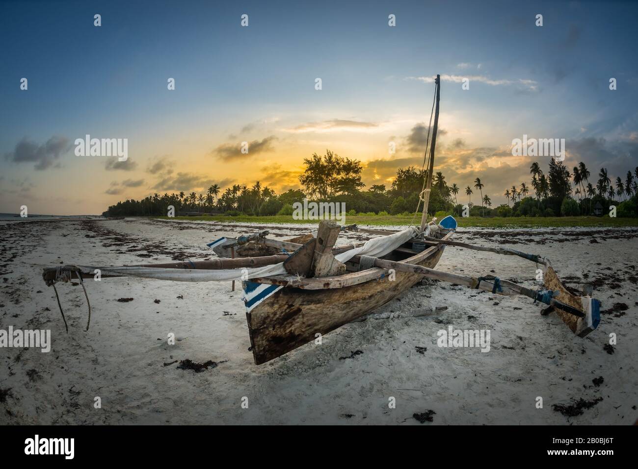 Traditionelles Holzboot bei Sonnenuntergang am tropischen Strand mit Palmen und weißem Sand am ...