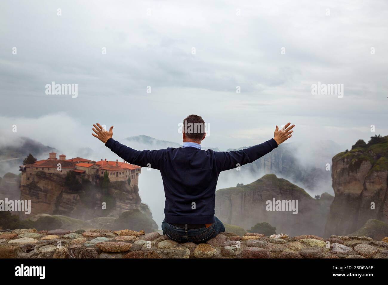 Der Mann sitzt an einer Wand und blickt auf das Kloster Meteora ...
