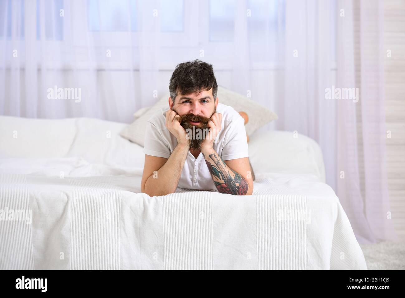 Kerl auf fröhlichem Gesicht, der auf dem Rand des Bettes auf weißen ...