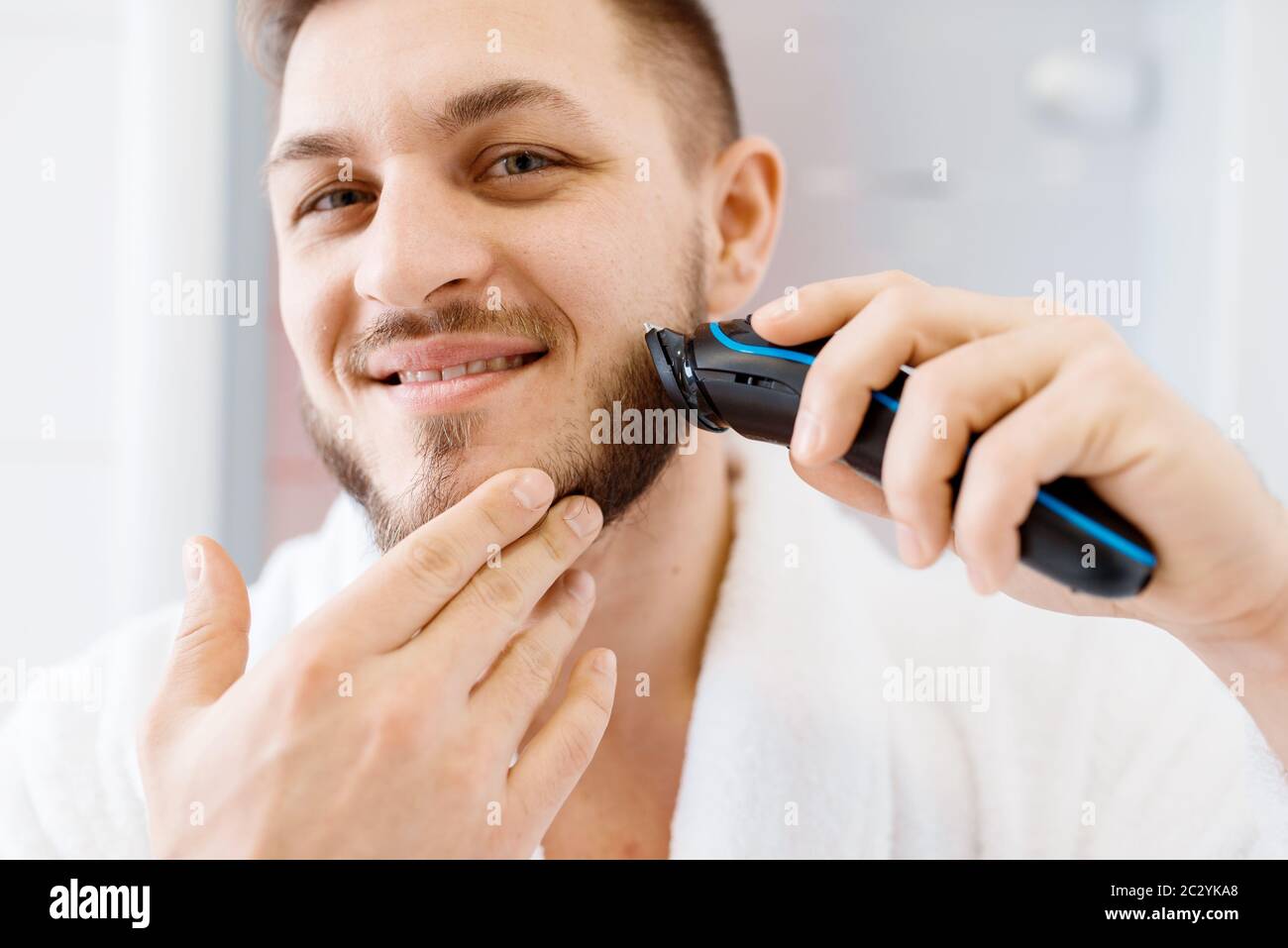 Mann im Bademantel rasiert seinen Bart mit einem elektrischen Rasierer im Bad, Routine ...