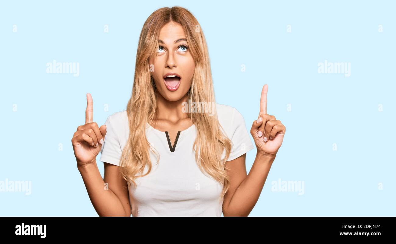 Schöne blonde junge Frau trägt beiläufige weiße T-Shirt erstaunt und ... Schöne blonde junge Frau trägt beiläufige weiße T-Shirt erstaunt und ...