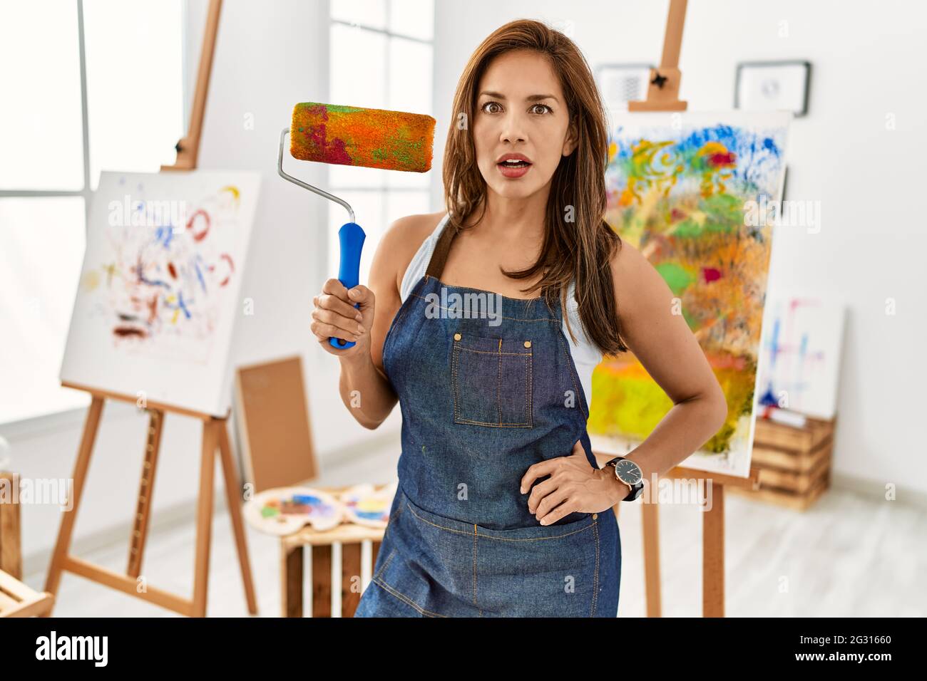 Junge lateinische Künstlerin Frau mit Malrolle, die im Kunststudio steht, erschrocken und ...