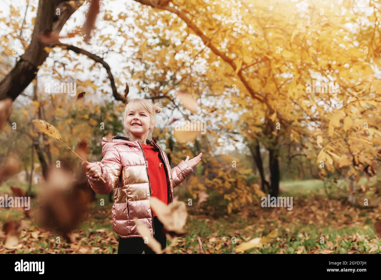 Glückliches kleines blondes Mädchen spielt mit gelben Herbstblättern im Garten. Das Kind lächelt ...