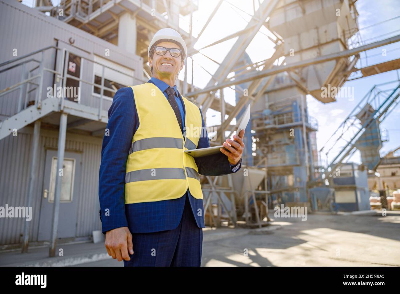 Fröhlicher männlicher Arbeiter mit Laptop, der im Werk im Freien steht ...