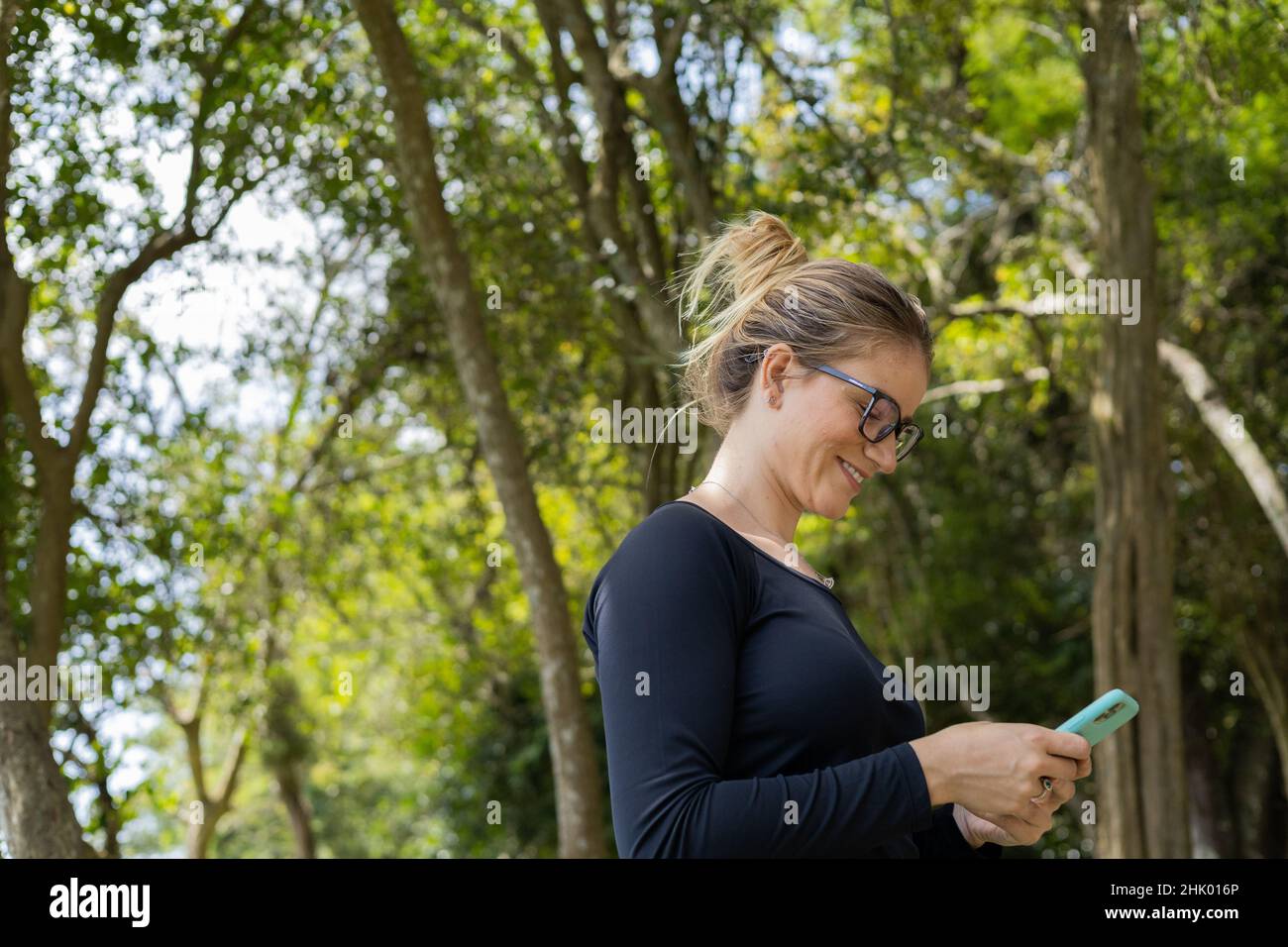 Die junge Frau benutzt tagsüber ein Smartphone mit einem grünen Park im ...