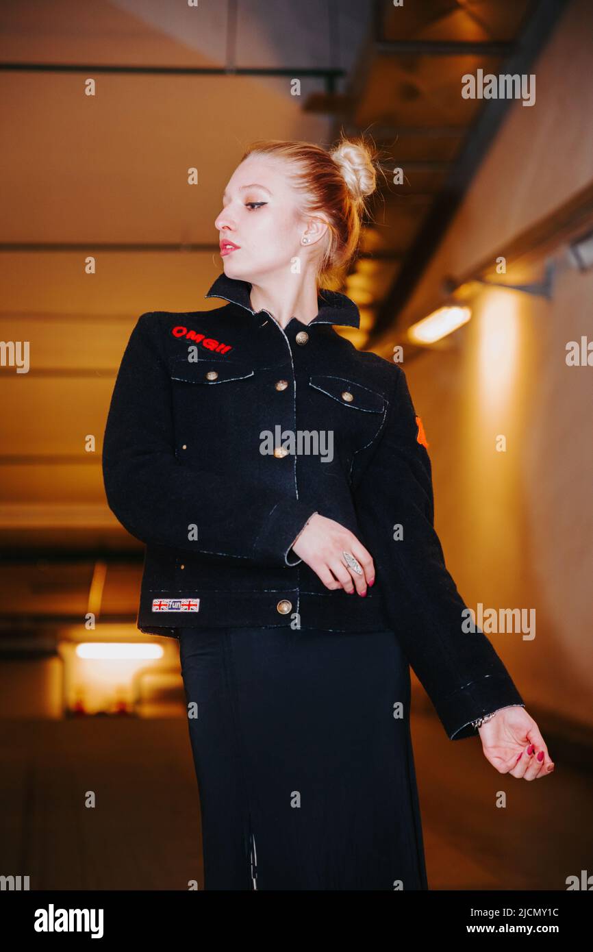 Das hübsche blonde Mädchen mit Haarsträhne, das einen langen schwarzen Mantel trägt, steht unter ...