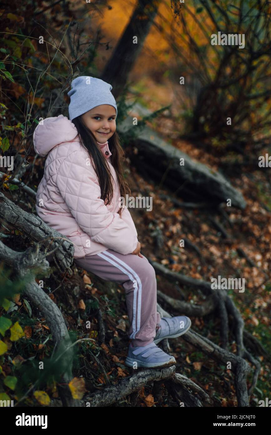 Junges kleines Mädchen, das auf Baumwurzeln im Wald sitzt, lächelt, die ... Junges kleines Mädchen, das auf Baumwurzeln im Wald sitzt, lächelt, die ...