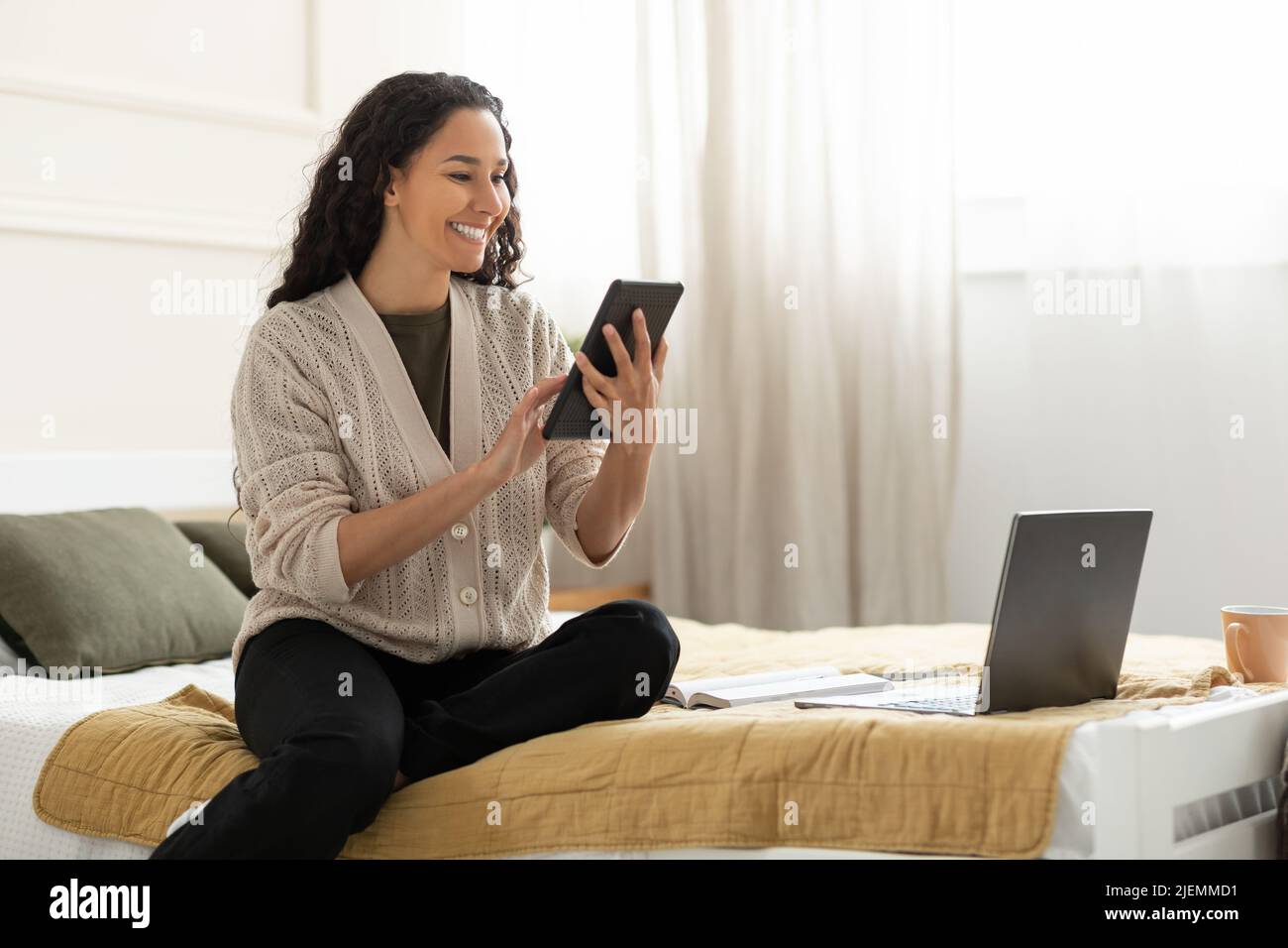 Schöne junge Frau, die auf dem Bett sitzt, zu Hause am digitalen Tablet ...