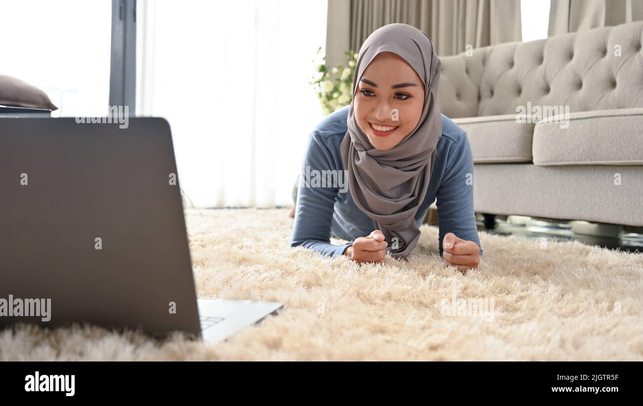 Aktive junge asiatische muslimische Frau mit Hijab, die im Wohnzimmer Übungen macht und über ...