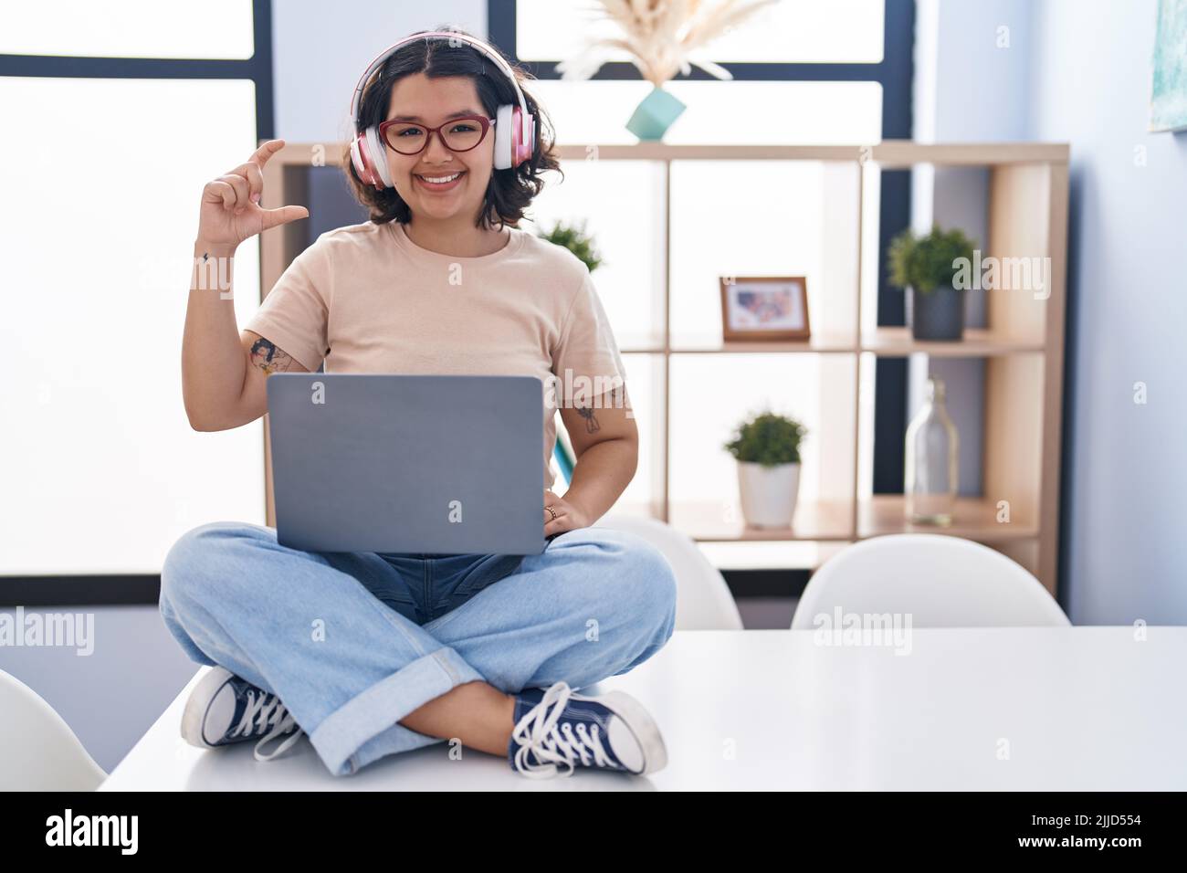 Junge hispanische Frau mit Laptop sitzt auf dem Tisch mit Kopfhörern lächelnd und selbstbewusst ...