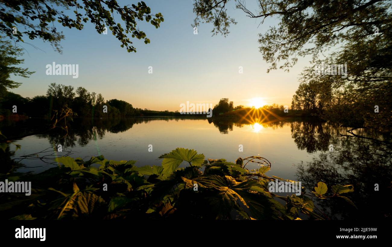 Der See mit Bäumen, die sich darin spiegeln, und die untergehende Sonne ...