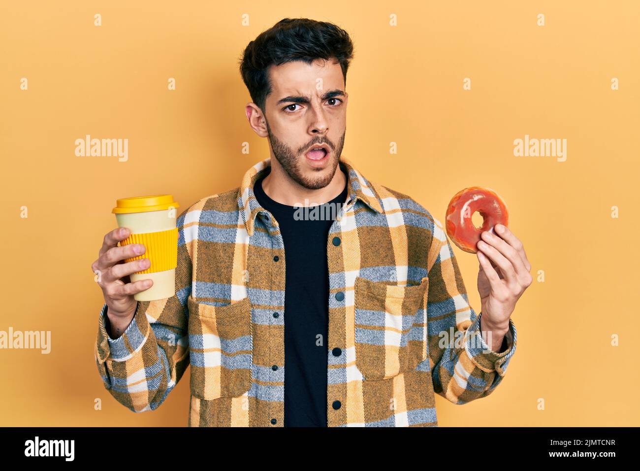 Junger hispanischer Mann, der Donut isst und Kaffee trinkt, schaut ... Junger hispanischer Mann, der Donut isst und Kaffee trinkt, schaut ...