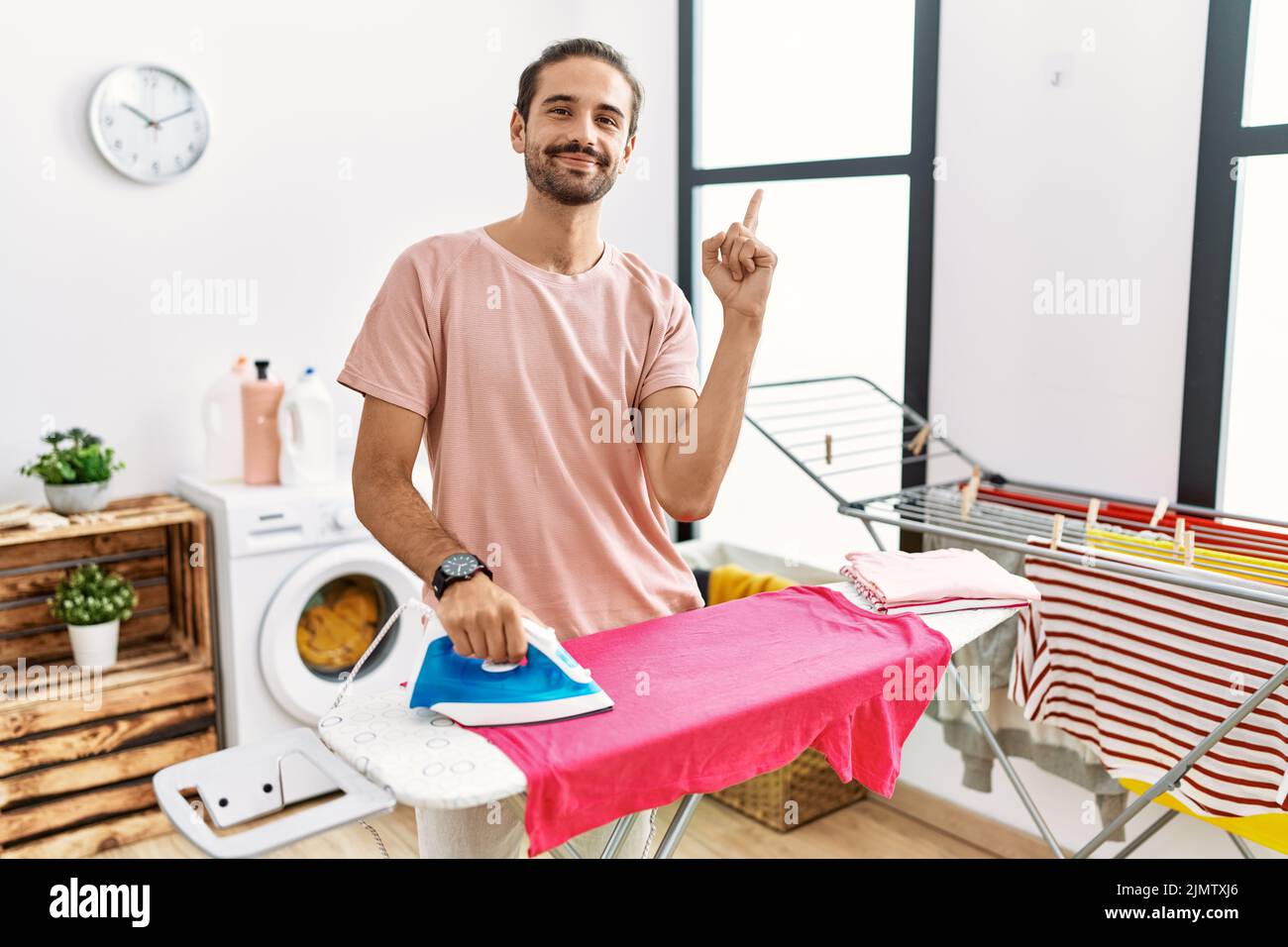 Der junge hispanic-Mann bügelt zu Hause mit einem großen Lächeln auf ...