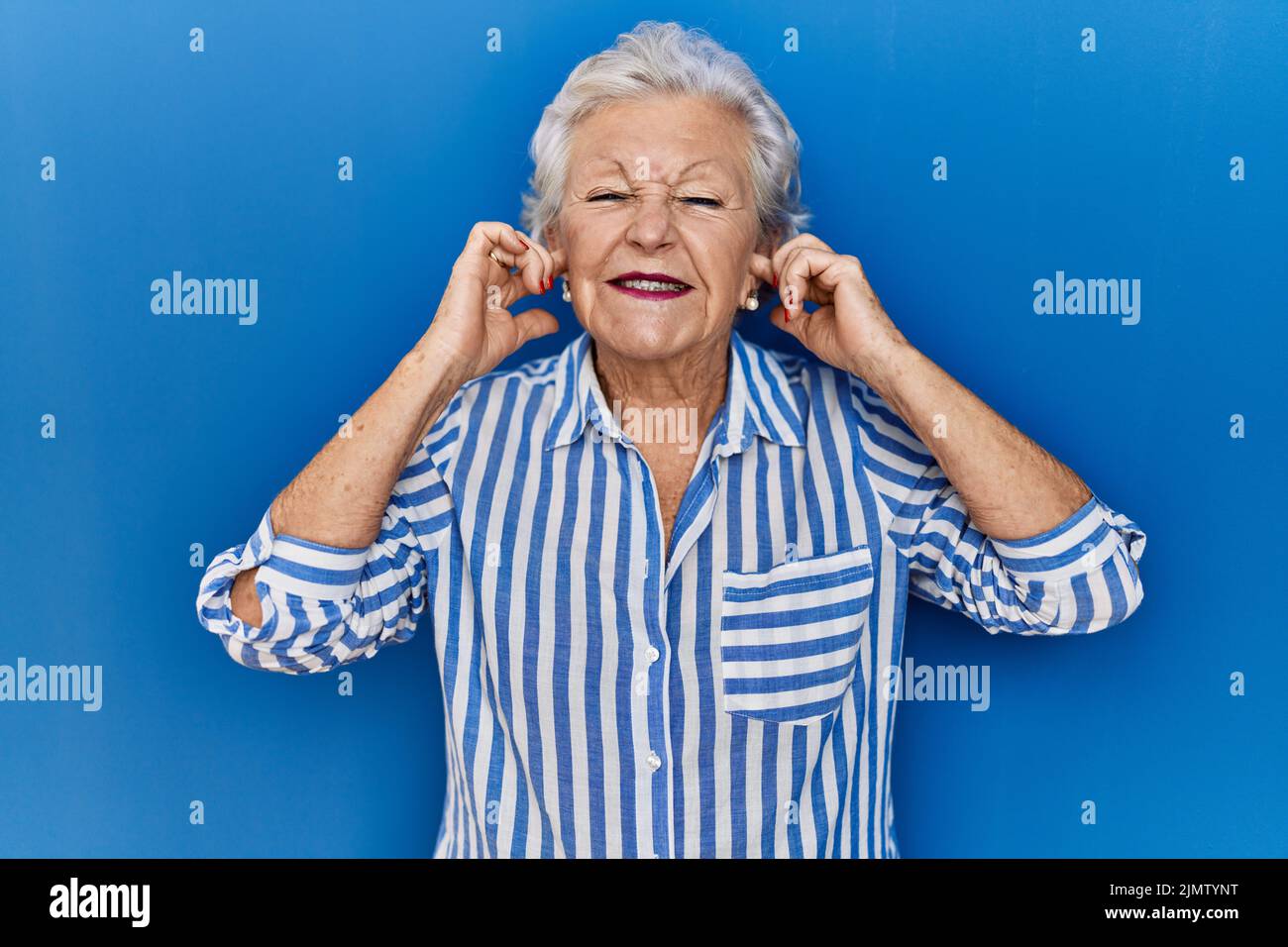 Ältere Frau mit grauen Haaren, die auf blauem Hintergrund die Ohren mit ...
