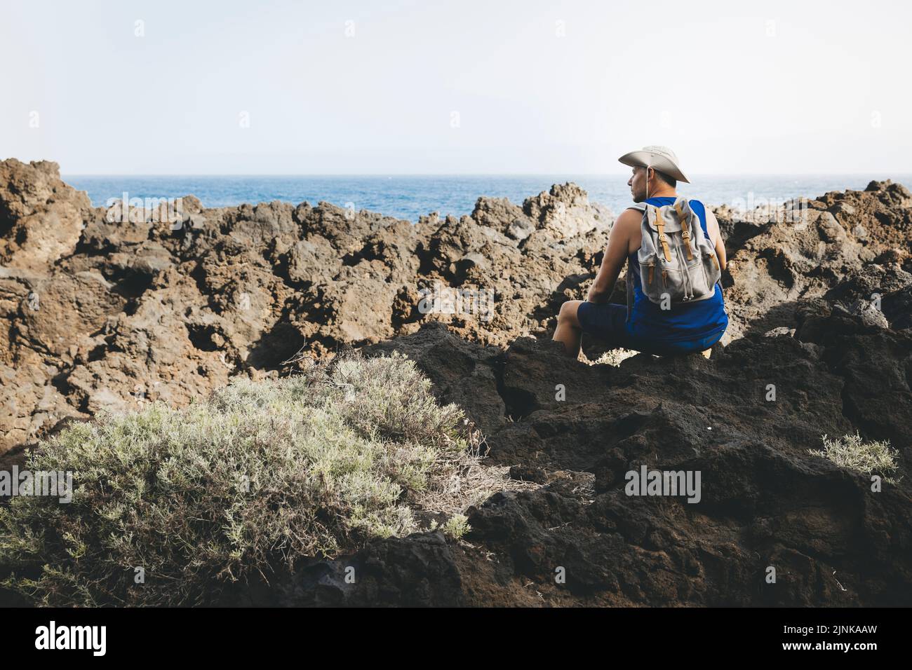 Wanderer mit Hut sitzt auf vulkanischem Felsen, hat Rucksack auf dem ...
