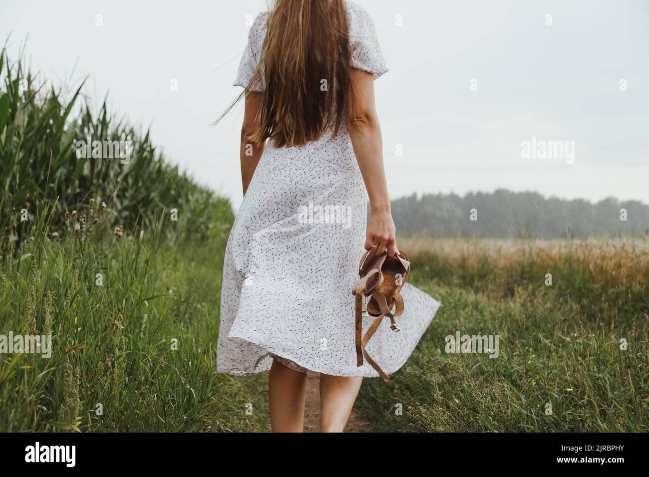 Die junge, fröhliche blonde Frau im nassen Sommerkleid mit Schuhen in den Händen läuft barfuß im ...