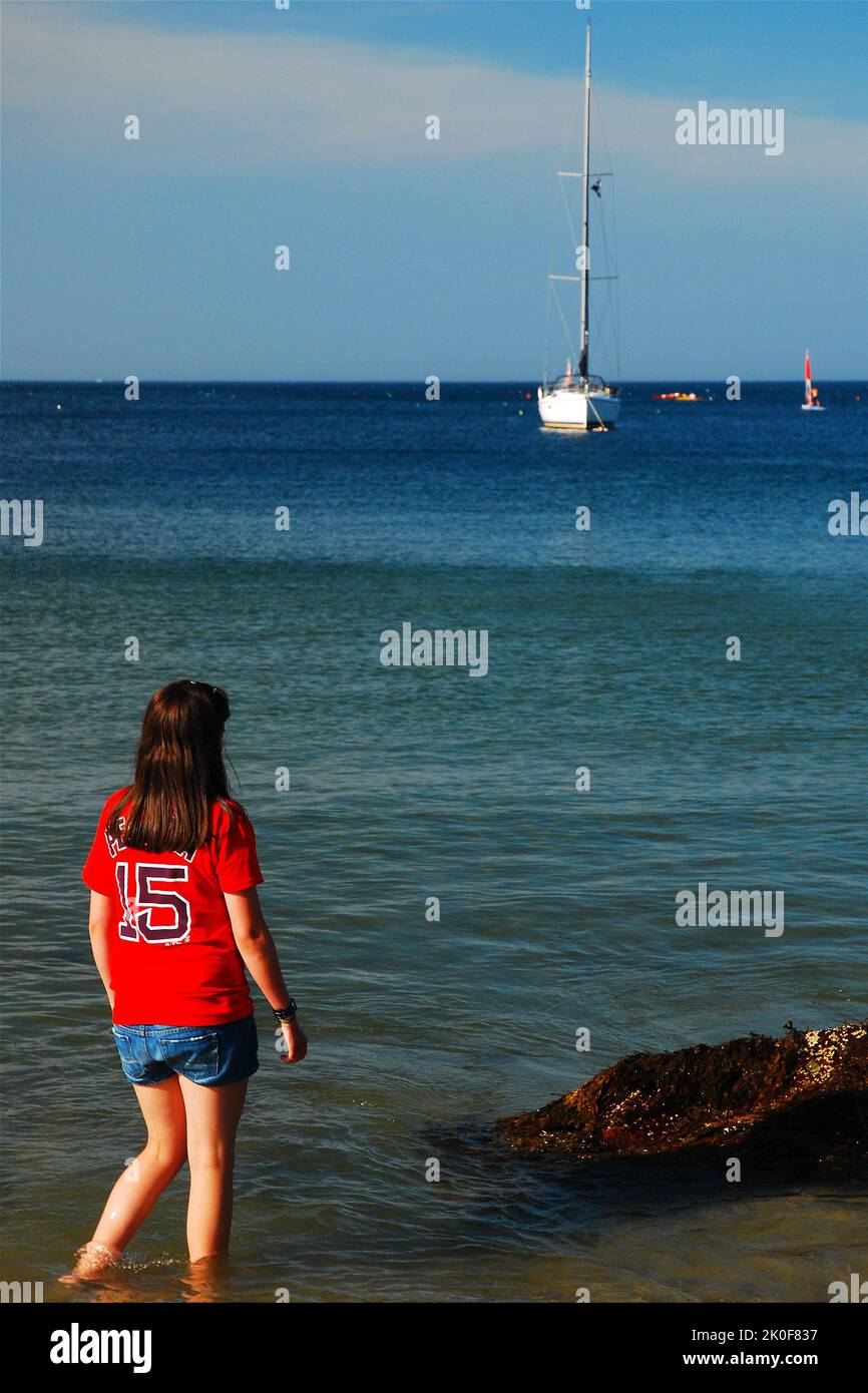 Eine junge Frau watet am Rand des Wassers und blickt auf das Meer auf das Segelboot, das an ...