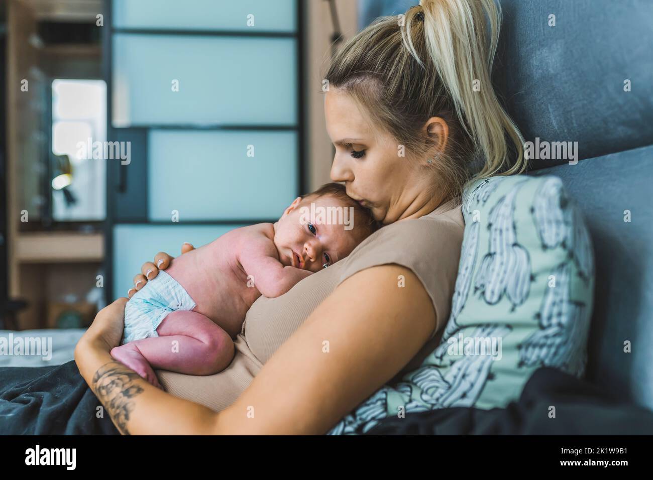 Weiße Frau mit blonden Haaren, die im Bett unter einer Decke ruht und ihren kleinen Jungen küsst ...