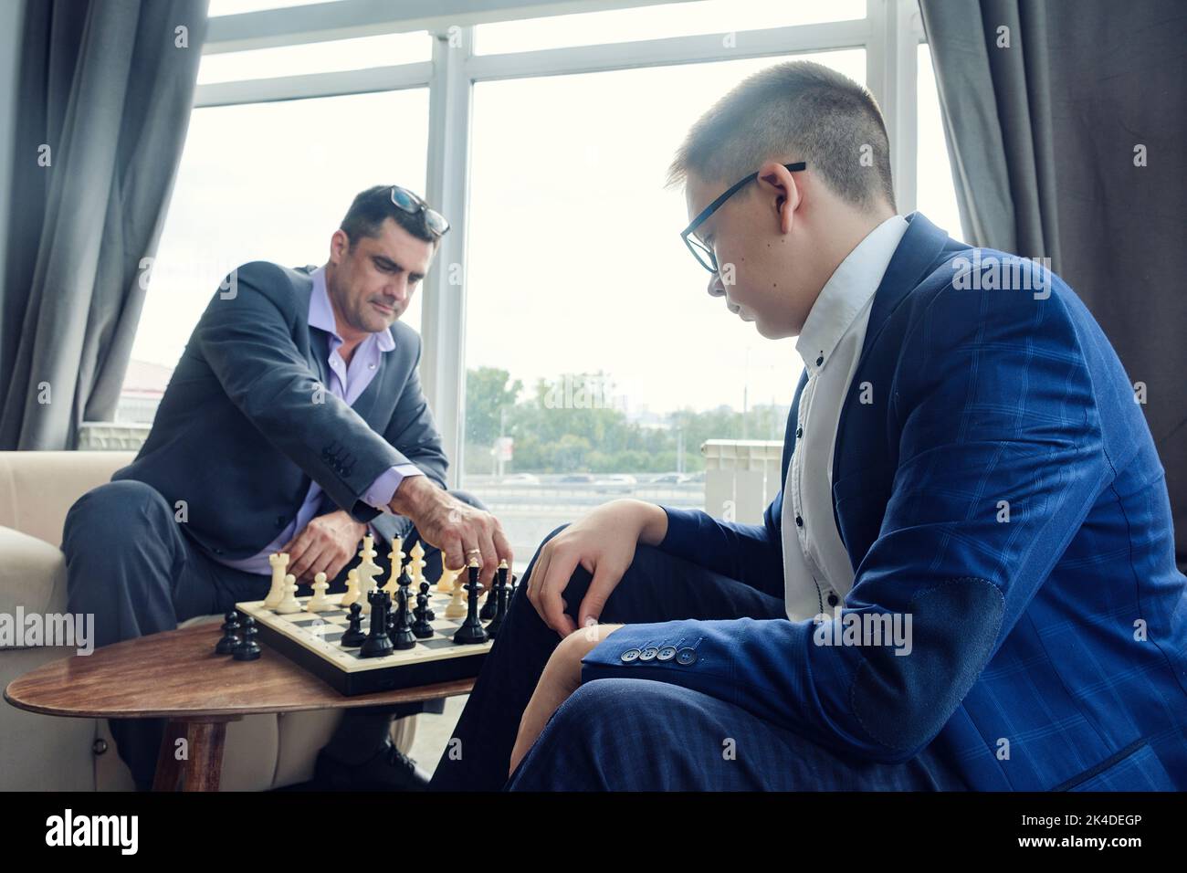 Vater und Sohn sitzen an einem Tisch an einem großen Fenster und spielen Schach und machen Züge ...