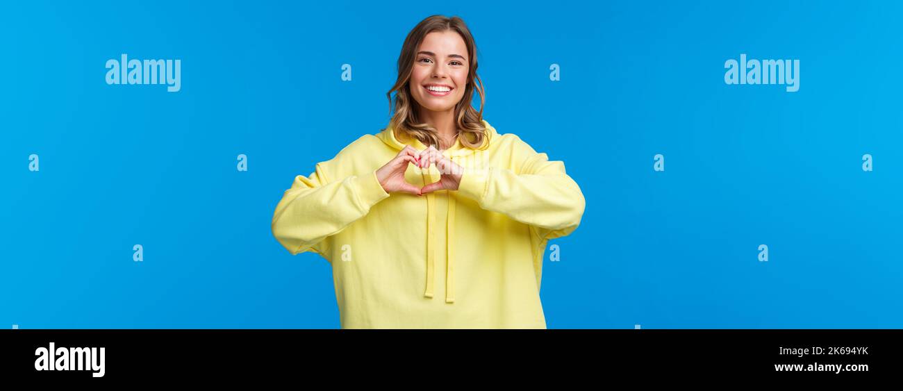 Liebenswürdige, süße blonde Mädchen in gelber Kapuze, die Herzgeste ...