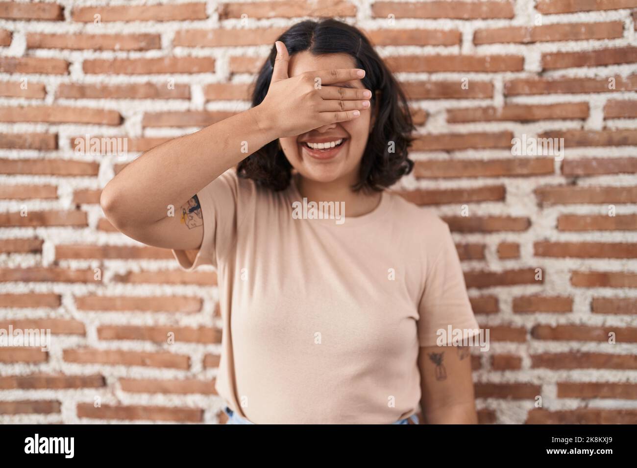 Junge hispanische Frau, die über einer Ziegelwand steht, lächelt und ... Junge hispanische Frau, die über einer Ziegelwand steht, lächelt und ...