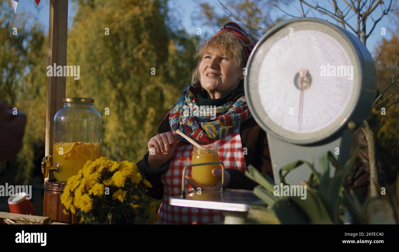 Ältere Verkäuferin steht am Stand und gibt Honig zum Geschmack. Die ...