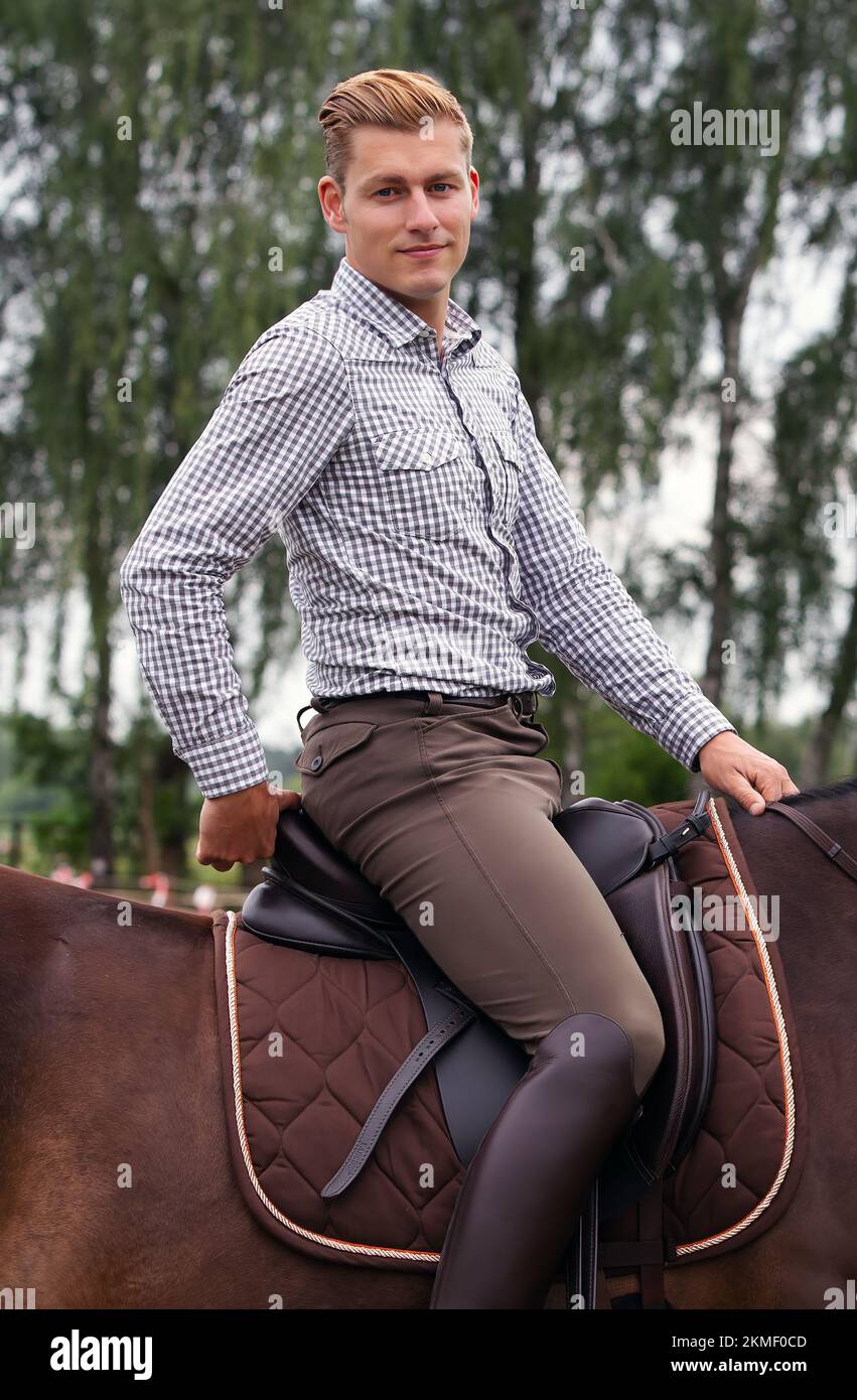 Hübscher junger Mann, der auf dem Land auf einem braunen Pferd reitet ...