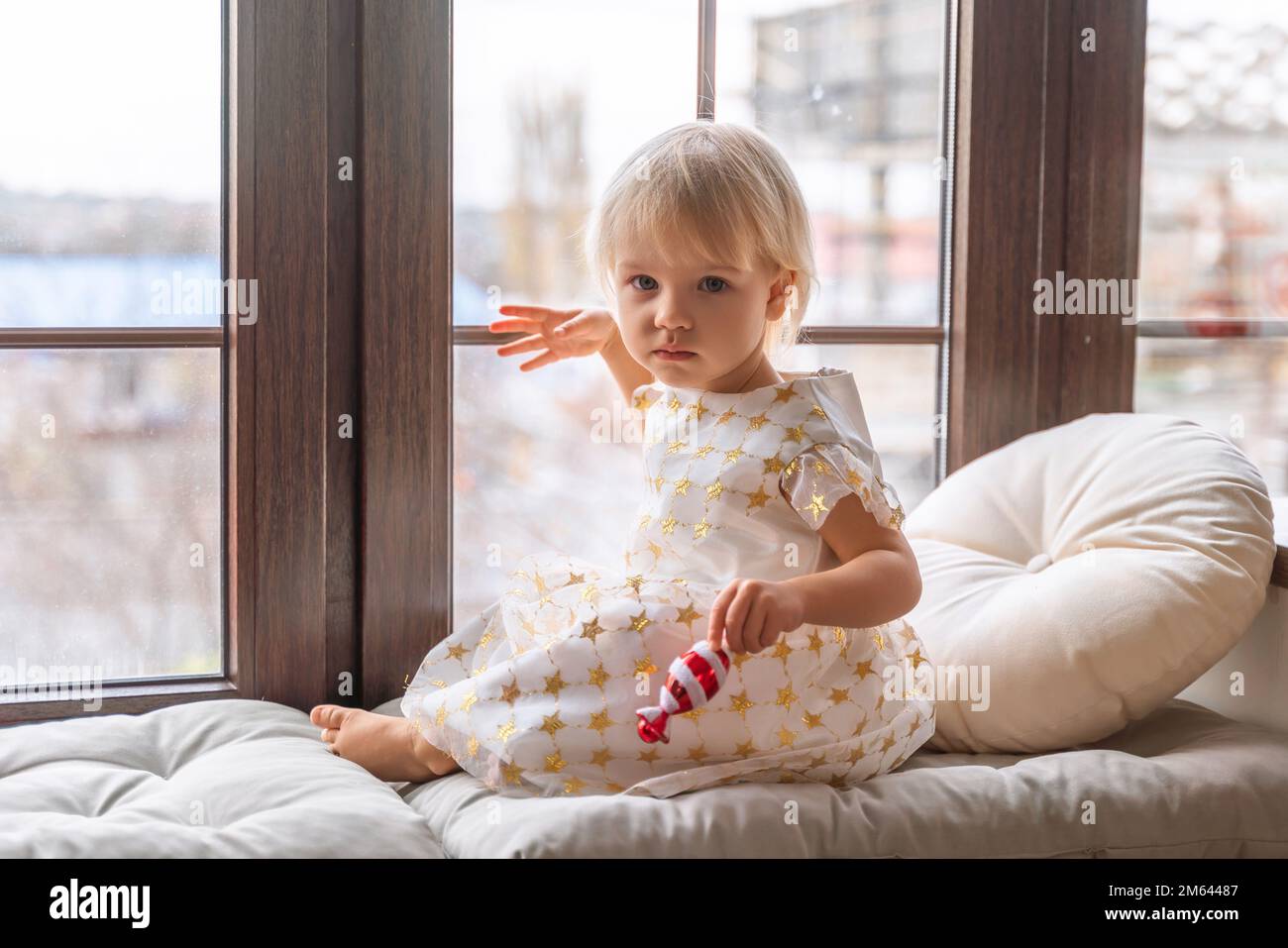 Mädchen mit 2 Jahren sitzt auf der Fensterbank. Ein kleines blondes Mädchen in einem weißen ...