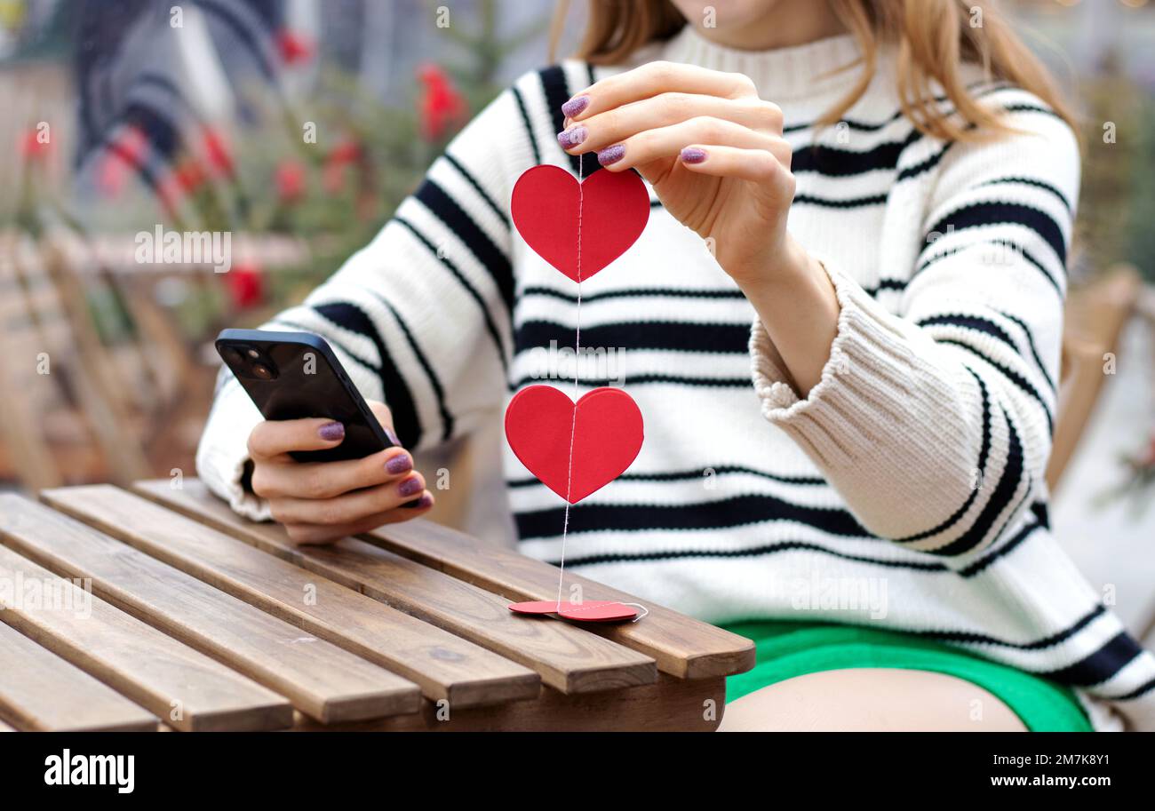 Frau sitzt auf dem Tisch mit Papierherzen Girlande in einer Hand und benutzt Smartphone mit ...