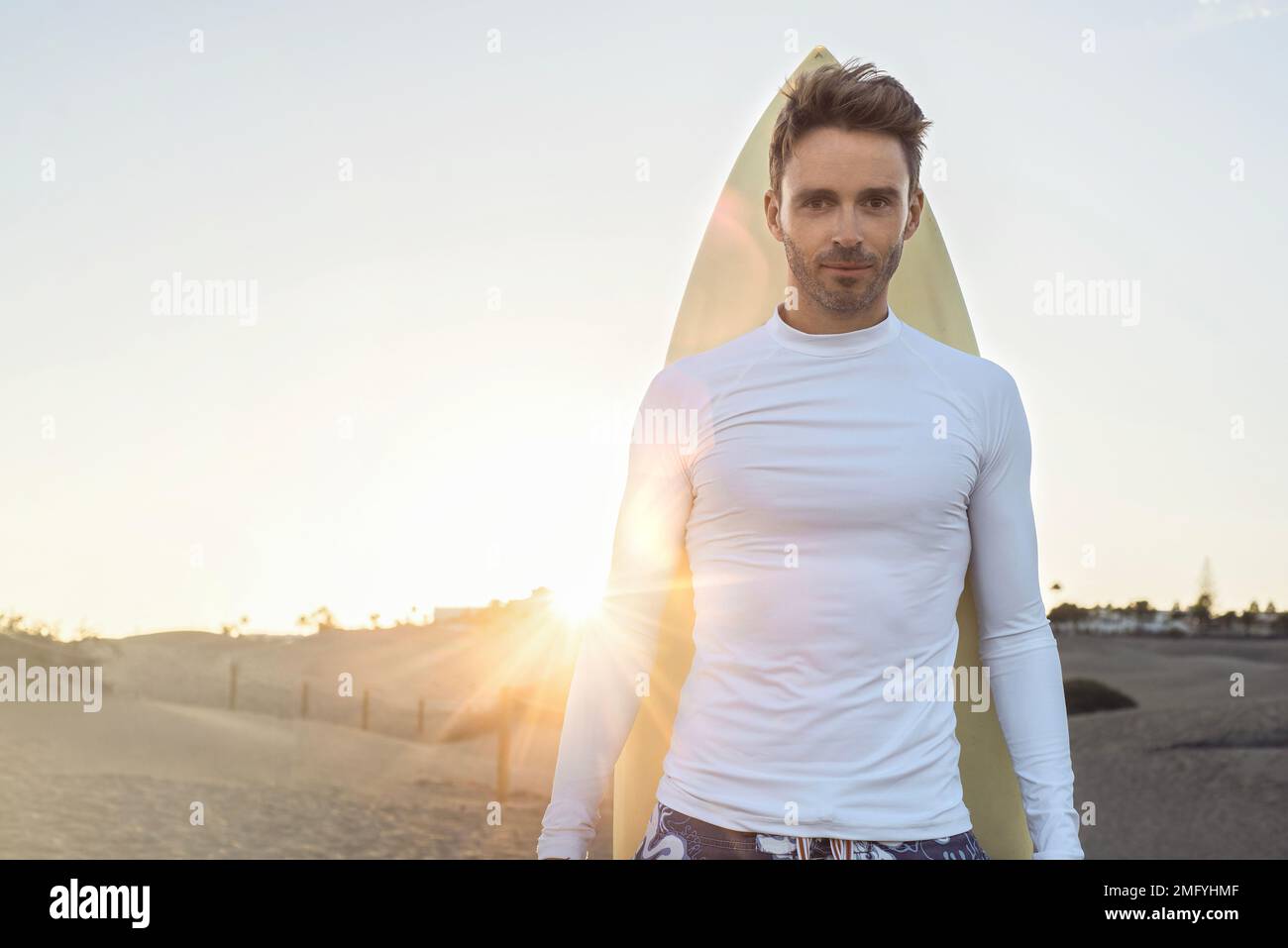 Der junge gutaussehende Surfer steht neben seinem Surfbrett und trägt ...