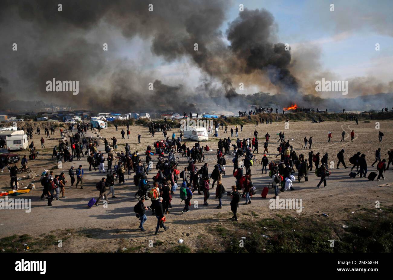 People walk past as thick smoke and flames rise from amidst the tents ...