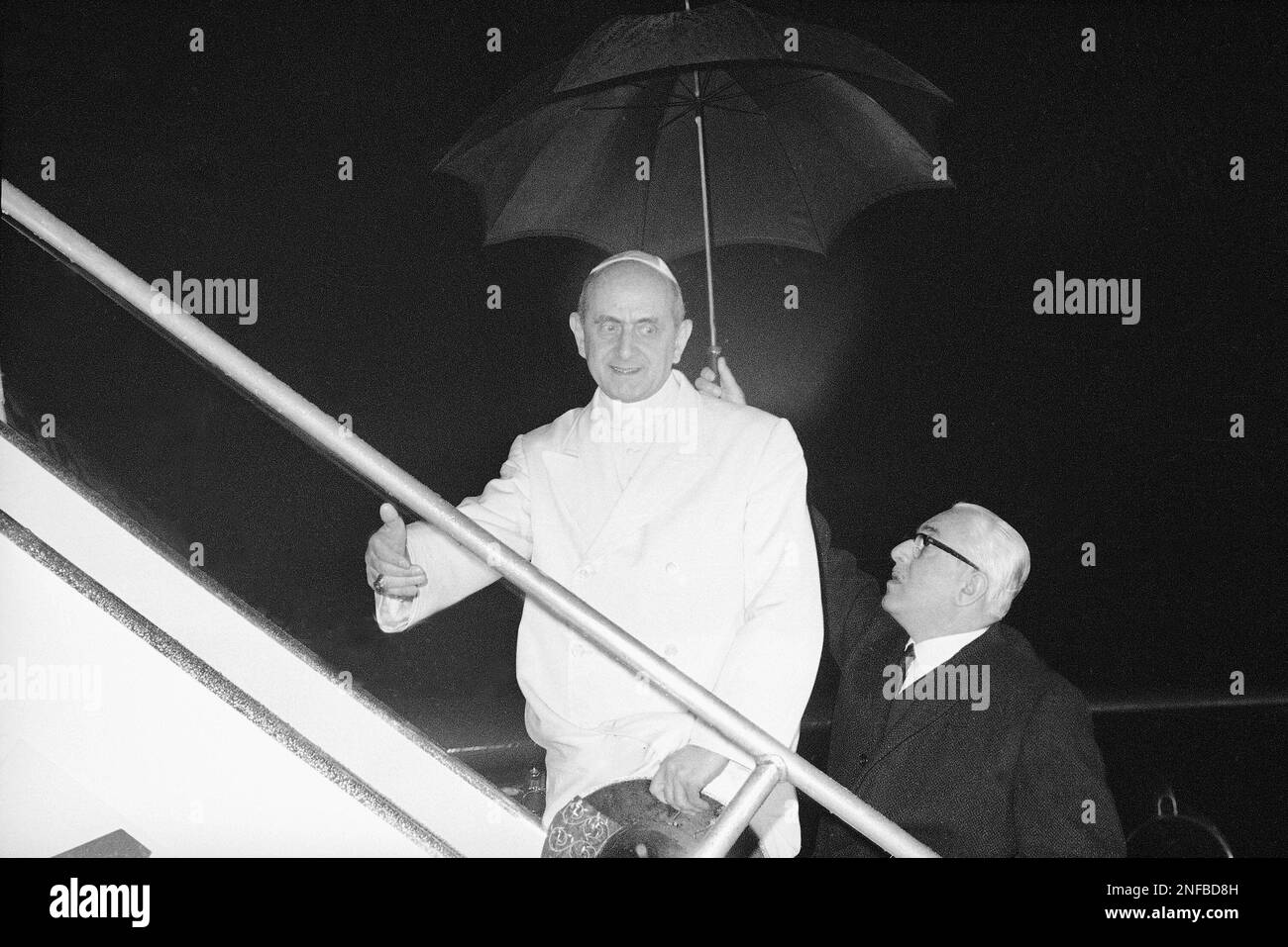 Pope Paul VI stands on a ramp to a plane under an umbrella held by ...
